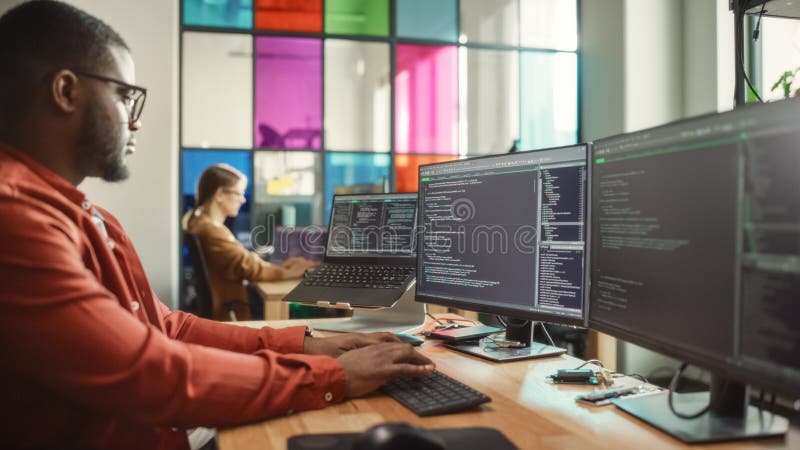 African American Man Writing Code on Desktop Computer with Multiple Monitors Set Up and a Laptop ...