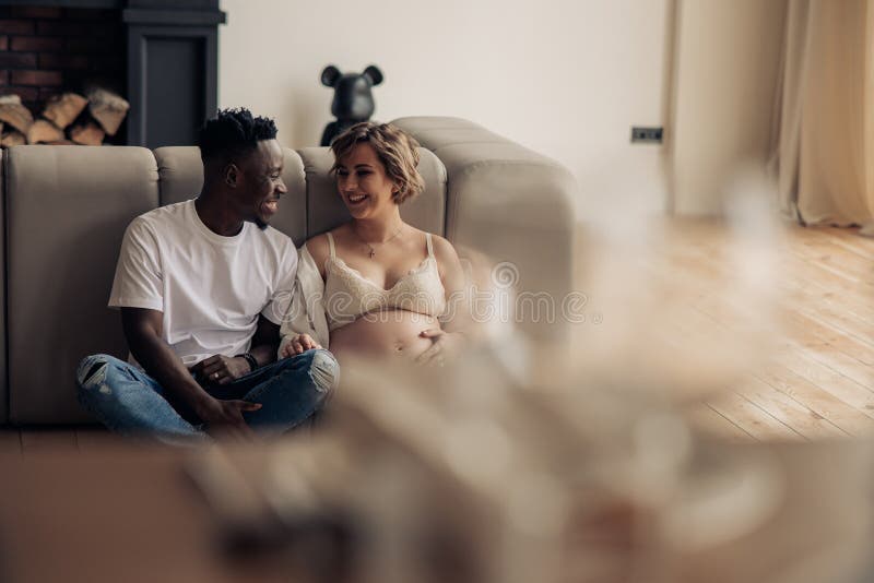 African Man And Caucasian Pregnant Woman Sit On Sofa And Laugh Joyfully