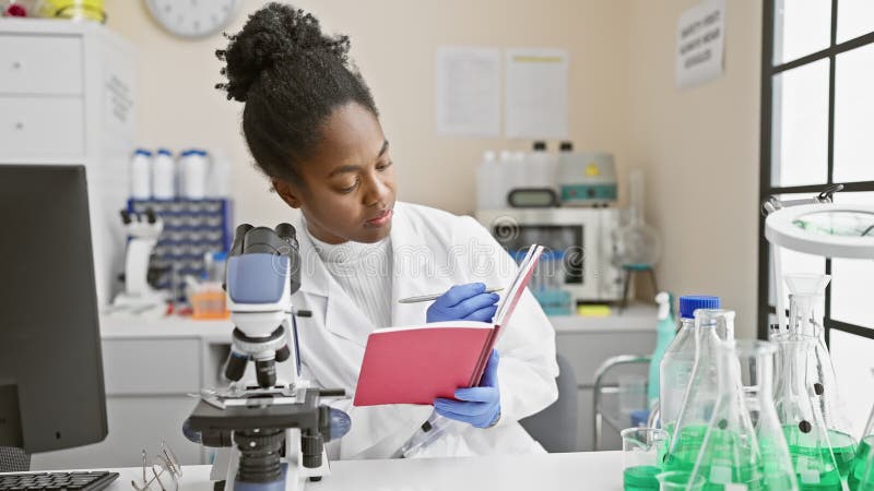 African Woman Scientist Observing through Microscope and Taking Notes ...