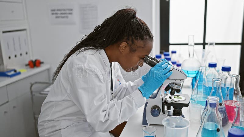 African Woman Scientist Using Microscope in Laboratory with Beakers ...