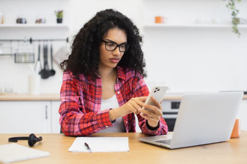 Focused Young Woman Using Smartphone while Working on Laptop in Kitchen ...