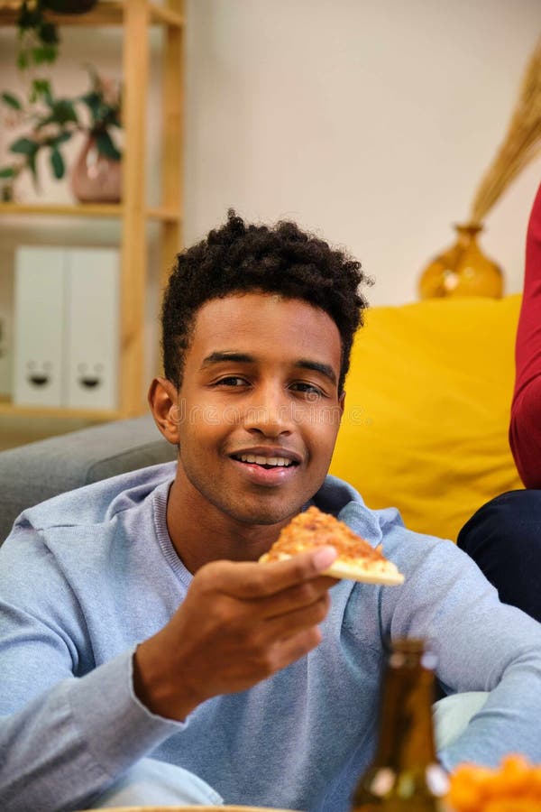 African Young Man Eating Pizza, Smiling and Looking at Camera at Home ...