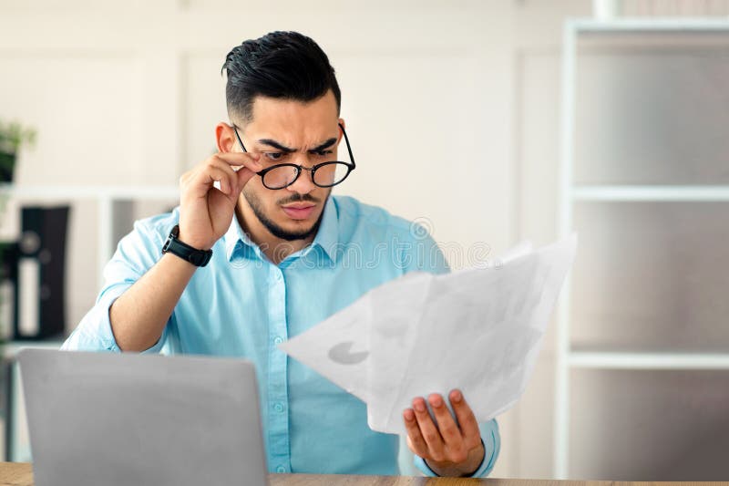Annoyed Arab Man Looking at Documents, Sitting at Desk with Laptop ...
