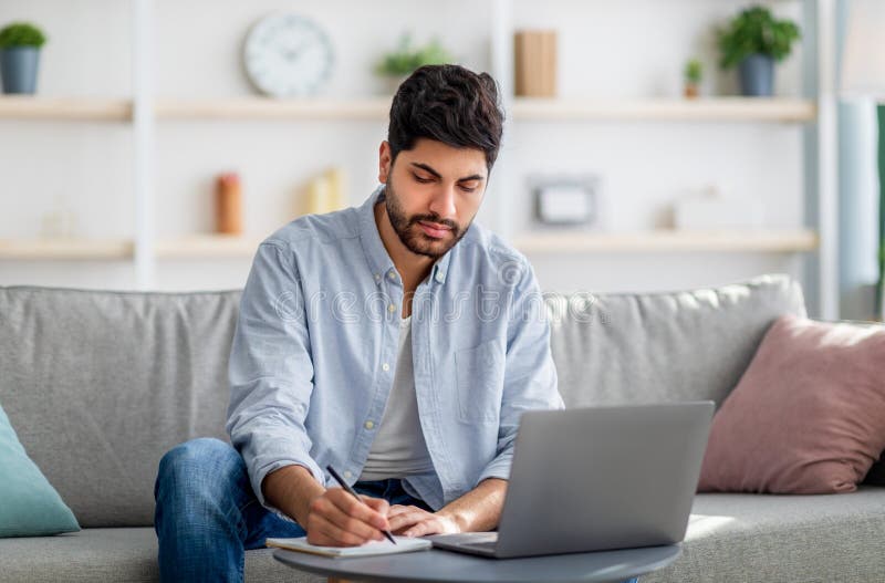 Arab Male Freelancer Writing in Notepad and Using Laptop, Taking Notes ...