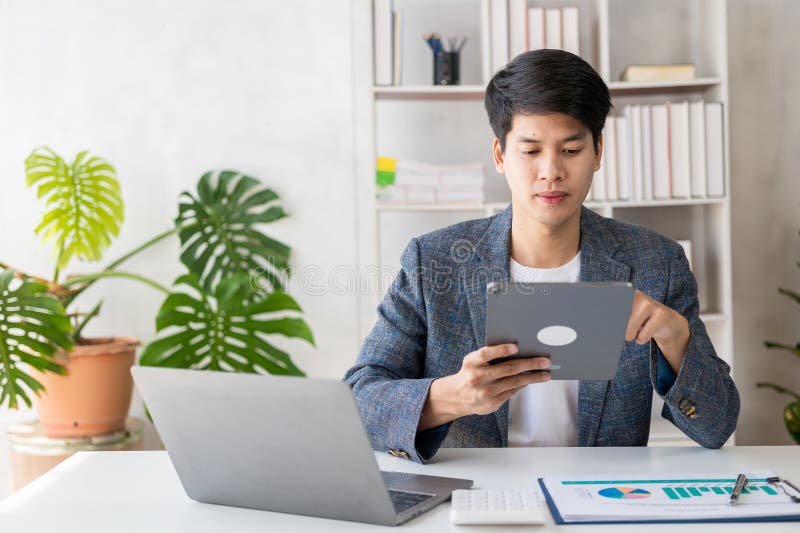 Asian Businessman Holding Tablet Working with Laptop and Financial Documents at Table in Office ...