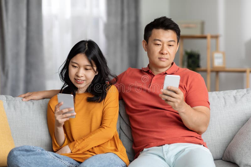 Asian Couple Sitting On Couch Together Using Their Smartphones Stock