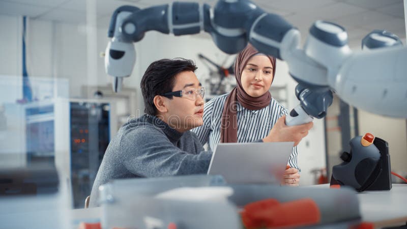 Asian Engineer Moving Robotic Arm with Control Panel and Developing Program on Laptop. Robotics ...