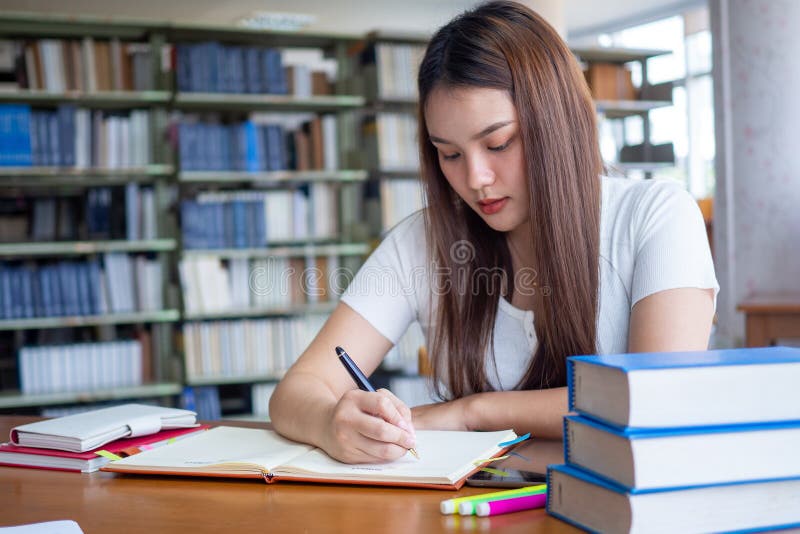 Asian Female Student Sitting at a Desk and Writing in a Notebook. Young ...