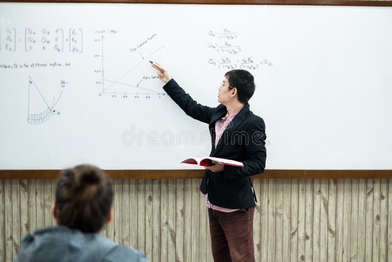An Asian Male Teacher in a Classroom Near the Whiteboard is Writing and ... An Asian Male Teacher in a Classroom Near the Whiteboard is Writing and ...