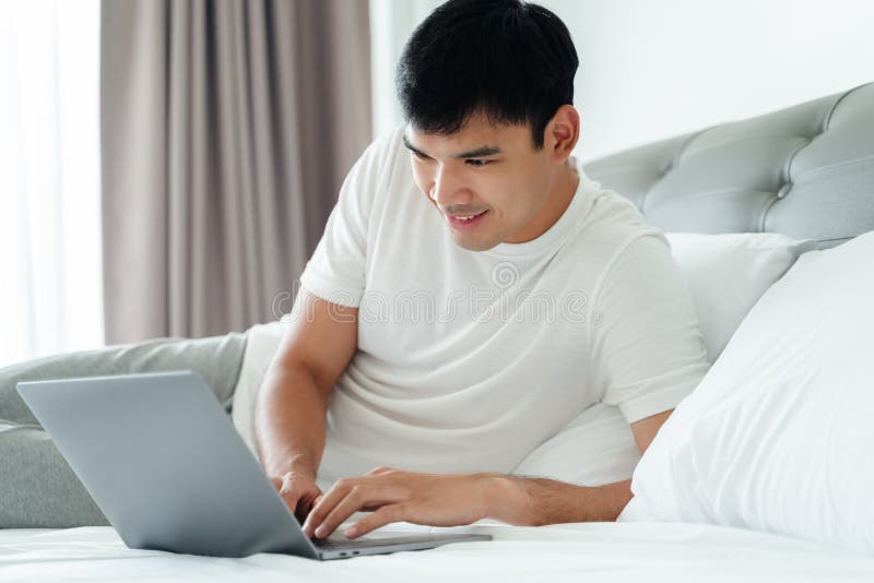 Asian Man in White T-shirt Laying on Bed Using Laptop Computer Having ...