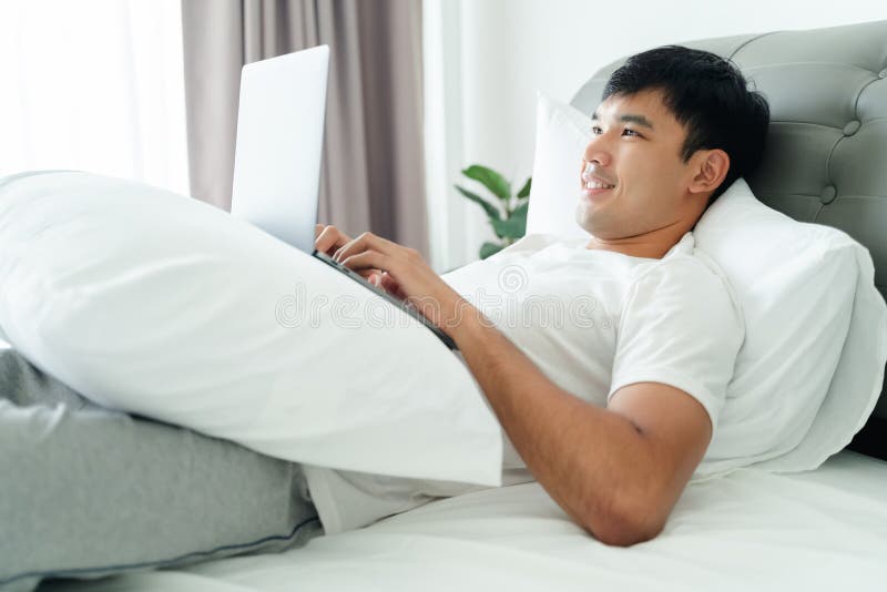 Asian Man in White T-shirt Laying on Bed Using Laptop Computer Having ...
