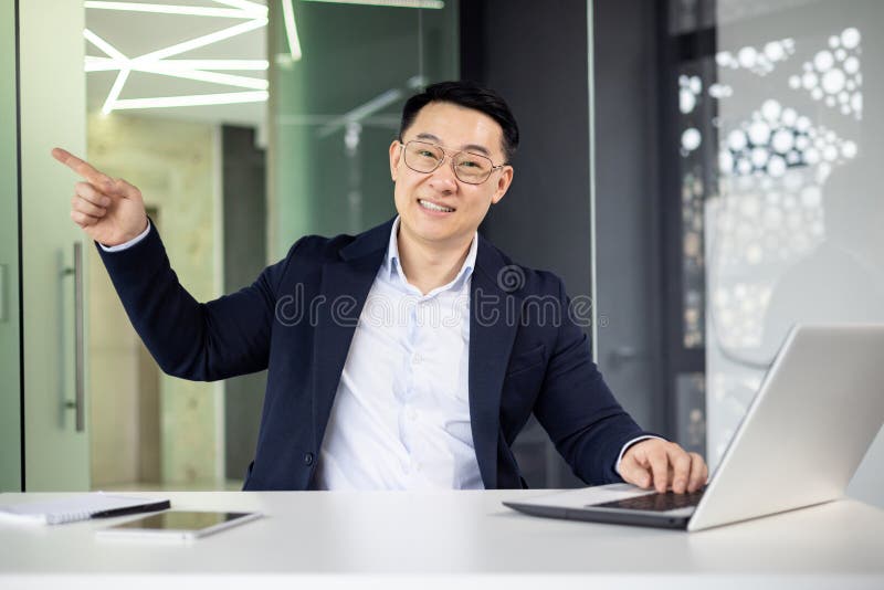 Asian Mature Man in Office Pointing Sideways while Working on a Laptop ...