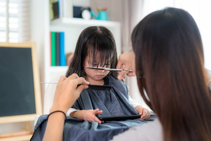 Asian Mother Cutting Hair To Her Daughter In Living Room At Home While