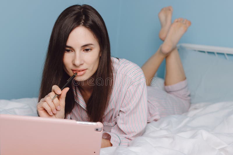 Attractive Serious Millennial Girl Lying In Bed Making Notes Watching Webinar Or Online
