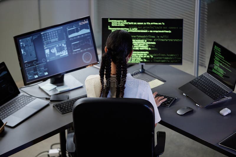 Back Of Female It Programmer Coding On Multiple Computers At Desk In