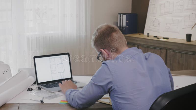 Back View of a Male Engineer Working on Laptop Computer with Blueprints ...
