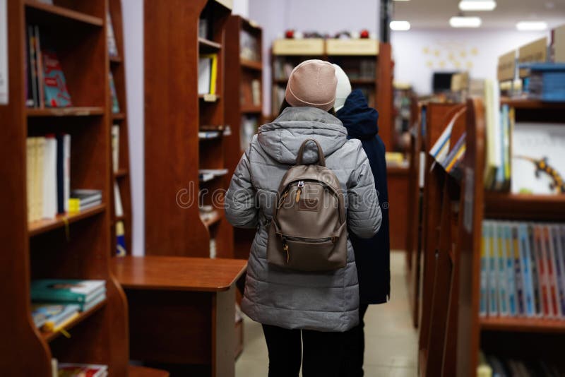 Back of Woman in Jacket and Backpack Reaching a Book from Bookshelf at ...
