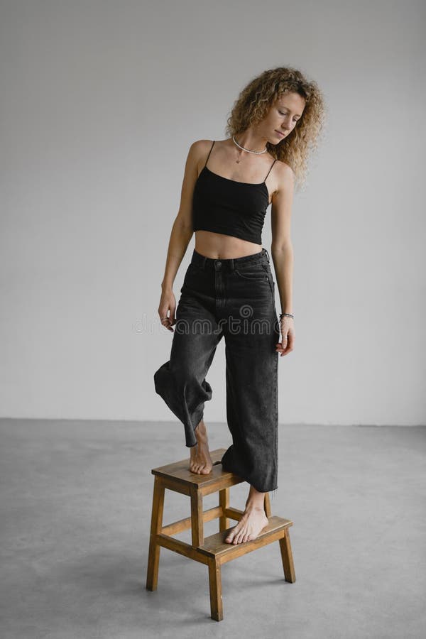 Barefoot and Curly Woman in Black Top and Jeans Standing on Step Stool ...