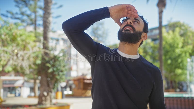 Bearded Man Shielding Eyes from Sun in an Urban Park Setting, Looking ...