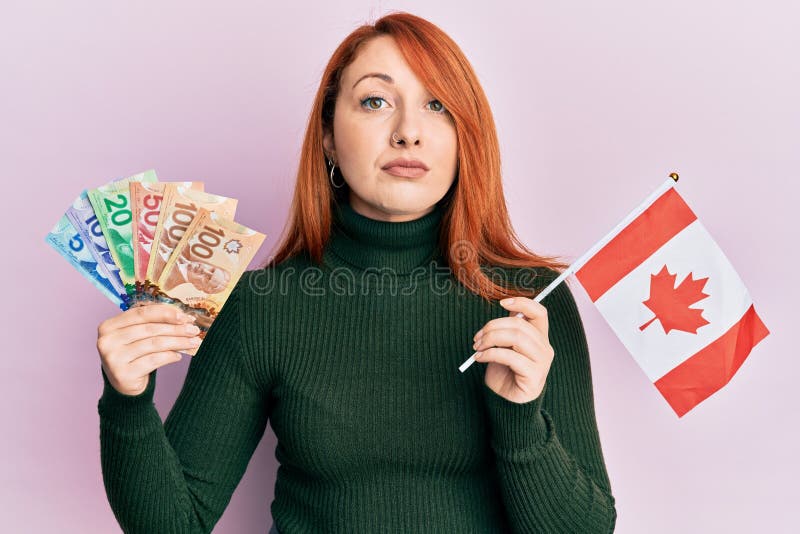 Beautiful Redhead Woman Holding Canadian Dollars And Canada Flag Relaxed With Serious Expression 5103