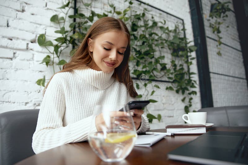 Beautiful Woman Sitting At The Table And Holding Mobile Phone Online