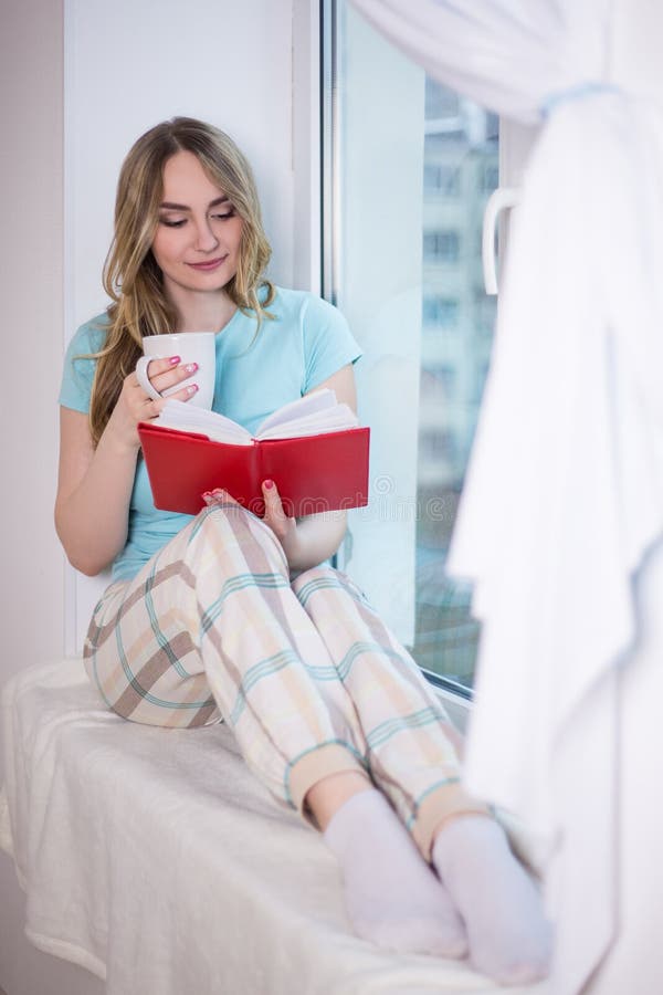 Beautiful Young Woman In Pajamas Sitting By The Window With Book Stock