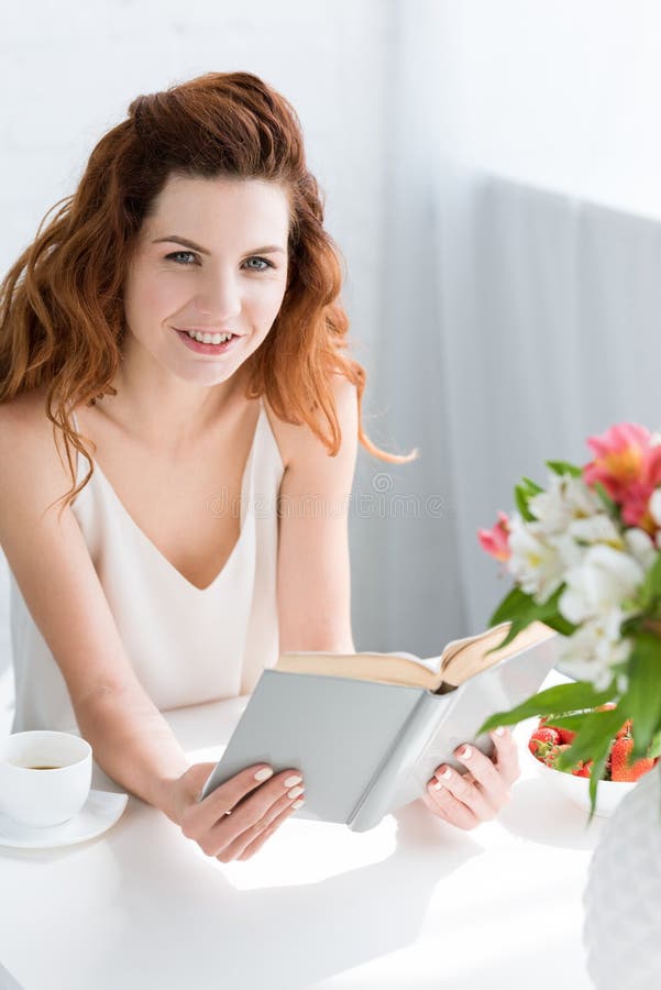 Beautiful Young Woman Reading Book While Sitting At Table With Coffee