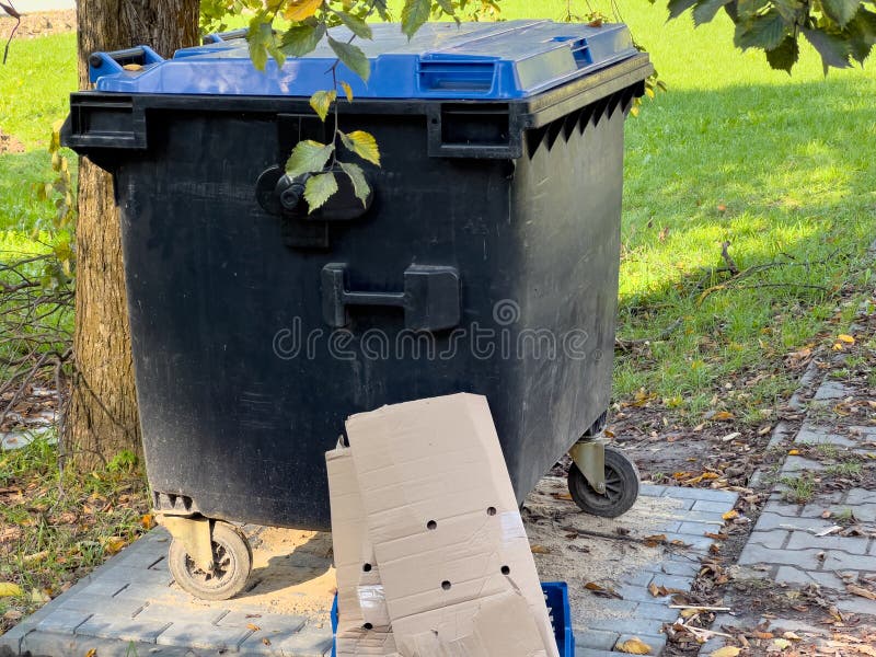 A Black and Blue Garbage Bin Sits on a Pathway Next To a Tree. a ...