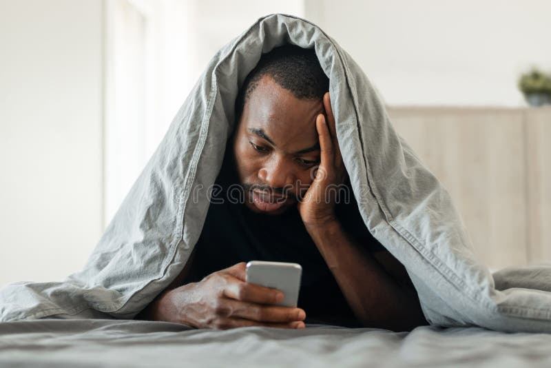Black Guy Using Smartphone Lying Covered with Blanket in Bedroom Stock ...