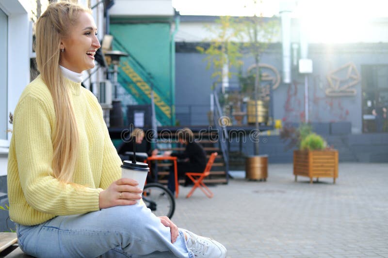 A Blonde Girl In A Yellow Sweater And Jeans Sits On A Bench With A Cup