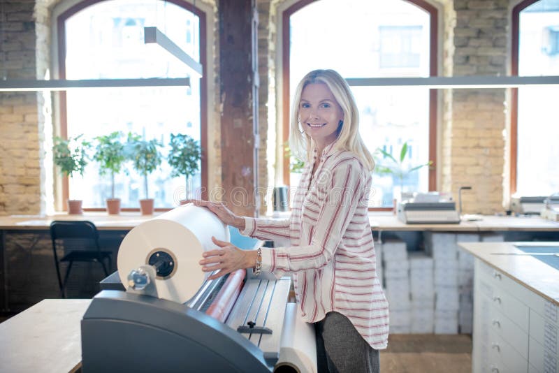 Blonde Woman Standing at Paper Roll Machine Next To the Window, Smiling ...