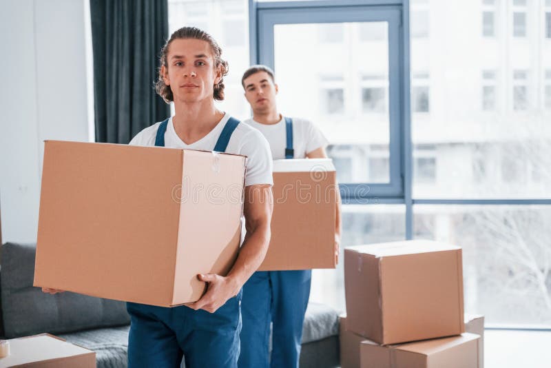 With Boxes in Hands. Two Young Movers in Blue Uniform Working Indoors ...