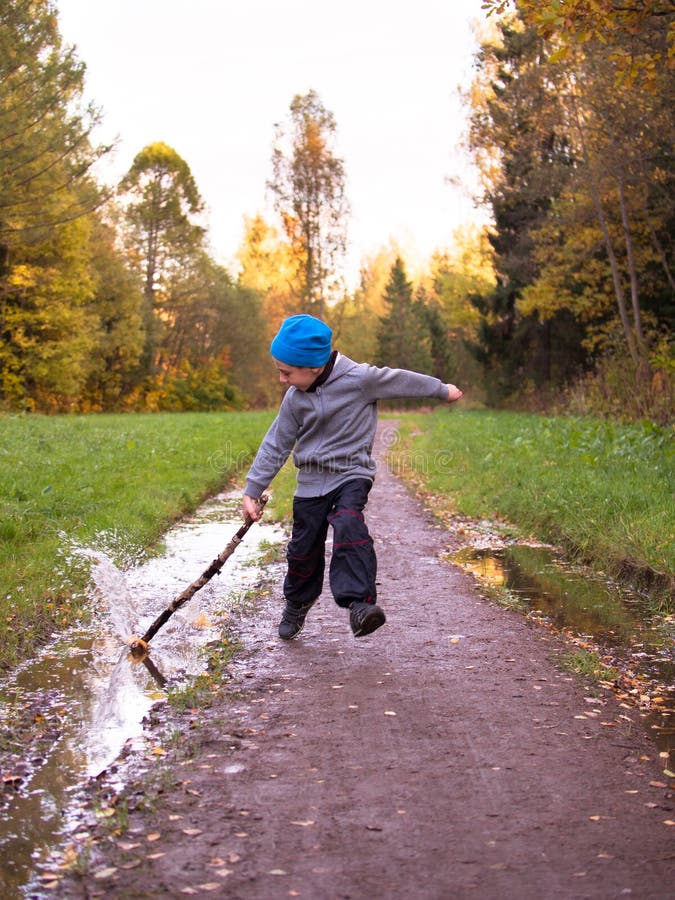 Boy in Hat Running Along the Path and Strikes Stick on Puddle Against ...