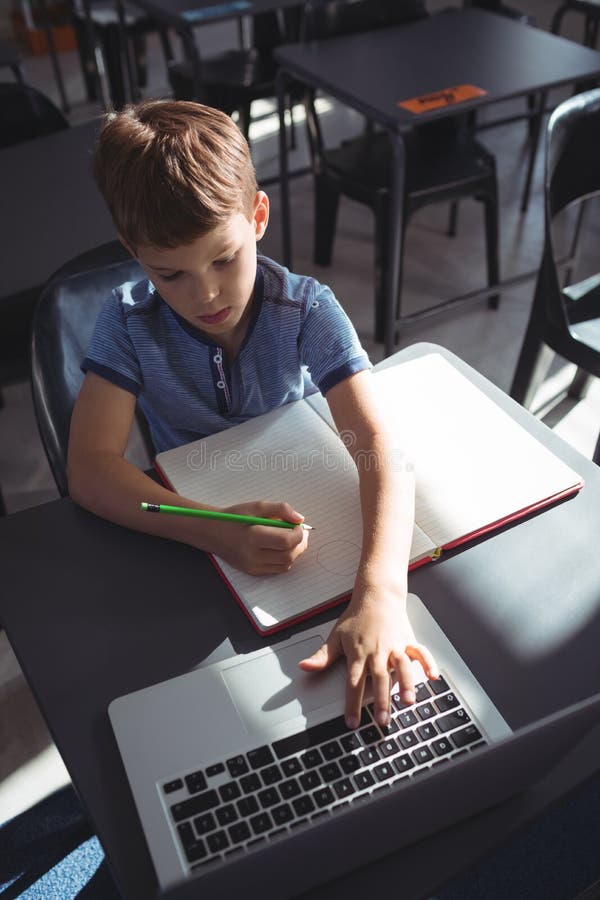 Boy Using Laptop while Writing in Book at School Stock Photo - Image of ...