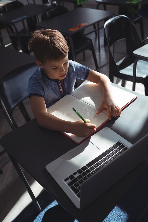 Boy Using Laptop while Writing in Book at School Stock Photo - Image of ...