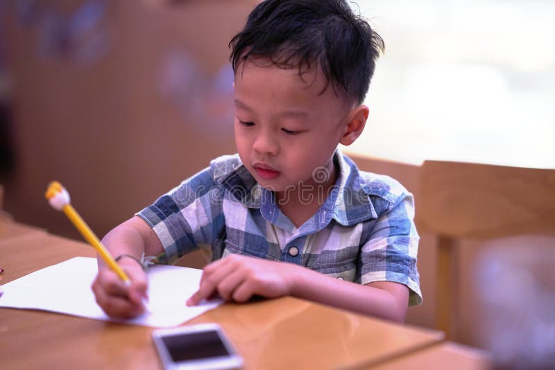 Boy is Writing Paper with Pencil and Parents`s Smart Phone on Table ...