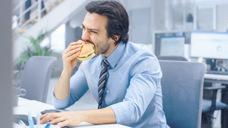 In the Bright Modern Office Young Businessman Eats Hamburger Sitting at ...