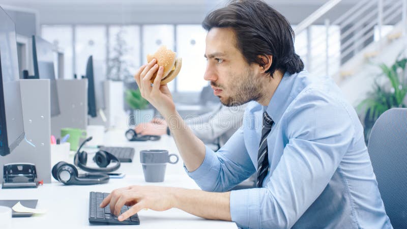 In the Bright Modern Office Young Businessman Eats Hamburger Sitting at ...