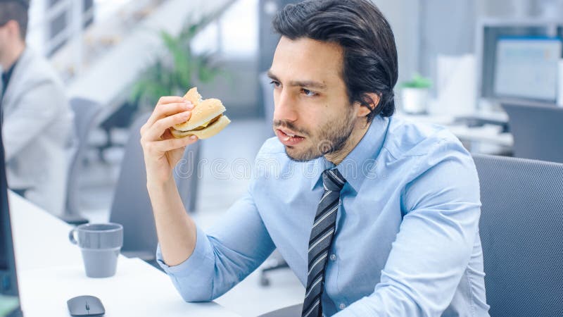 In the Bright Modern Office Young Businessman Eats Hamburger Sitting at ...