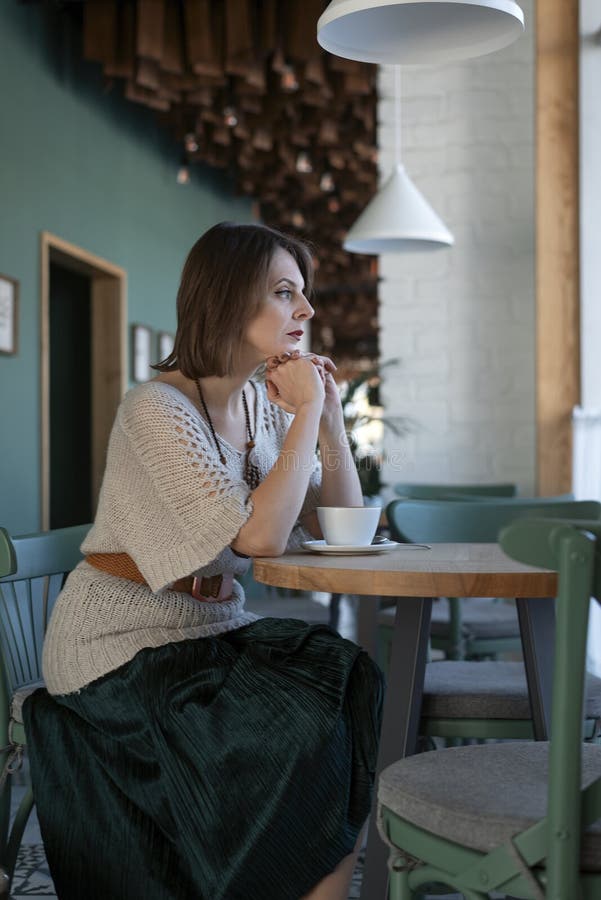 Brooding Young Woman Sits at Table in an Empty Cafe with Cup of Coffee ...