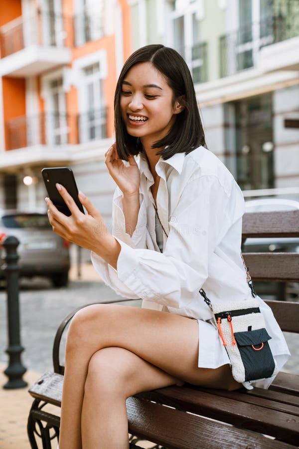 Brunette Asian Woman Smiling and Using Cellphone while Sitting on Bench Stock Image - Image of ...