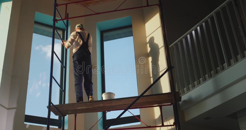 A Builder in Overalls Primes the Wall in the House. Stands on High ...