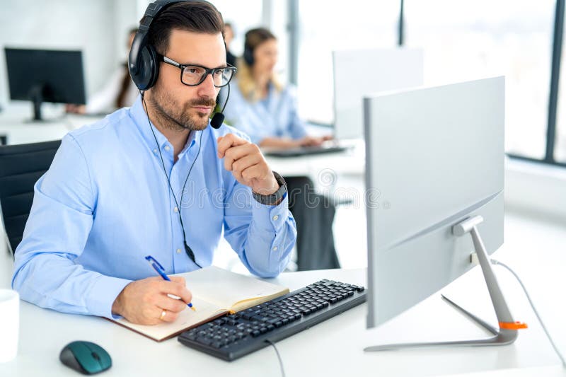 Business Man Wearing Headset Looking at Computer and Taking Notes To ...