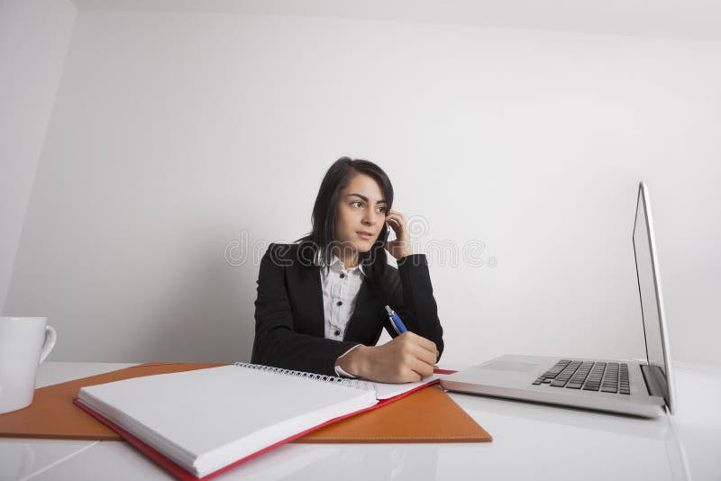 Businesswomen Using Cell Phone while Writing Notes from Laptop at ...