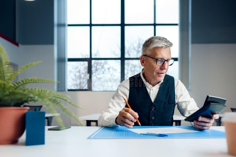 Busy Senior Man Sitting at Office Desk with Papers and Making Notes ...