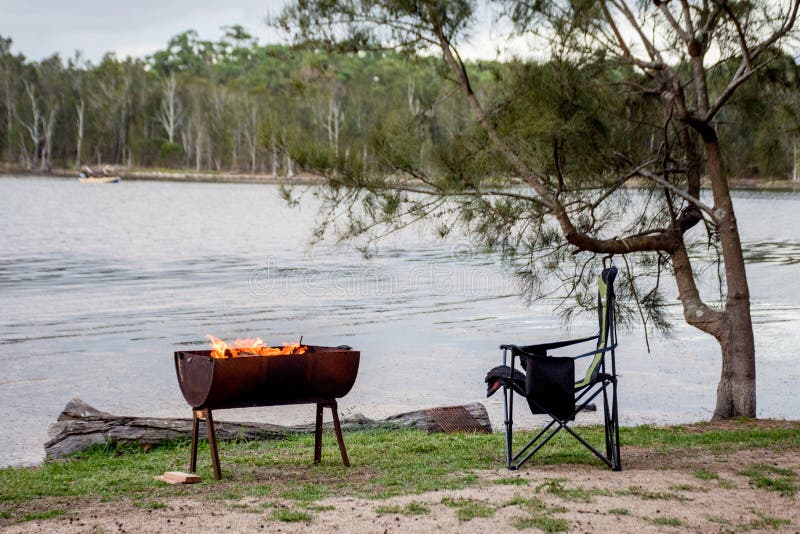 Campfire in a Half Barrel Fire Pit and Empty Campchair on the Lake ...