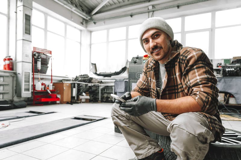 Car Service Worker Sitting on a Wheel and Resting, Using Mobile Phone ...
