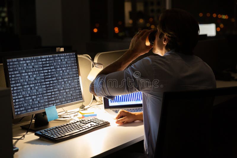 Caucasian Male Programmer Sitting At Desk Drinking Coffee Using Computer With Coding On Screen
