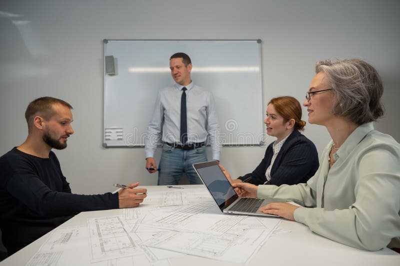 Caucasian Man Draws a Graph on a White Board, Three Colleagues Sit at ... Caucasian Man Draws a Graph on a White Board, Three Colleagues Sit at ...