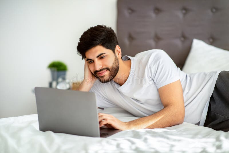 Cheerful Arab Man Browsing Internet on Laptop while Lying on Bed at ...